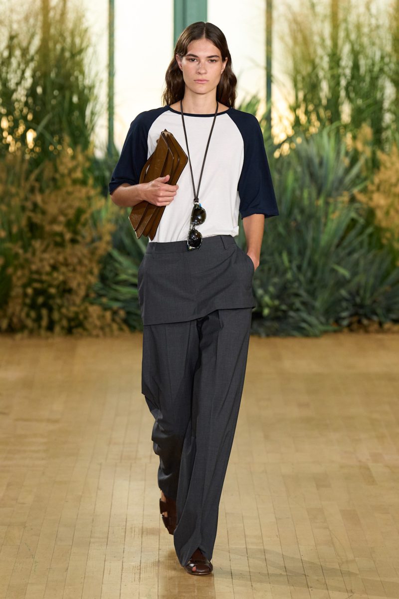 Model wearing a white and navy raglan sleeve top paired with high-waisted gray wide-leg trousers and carrying a brown leather clutch with sunglasses accessory