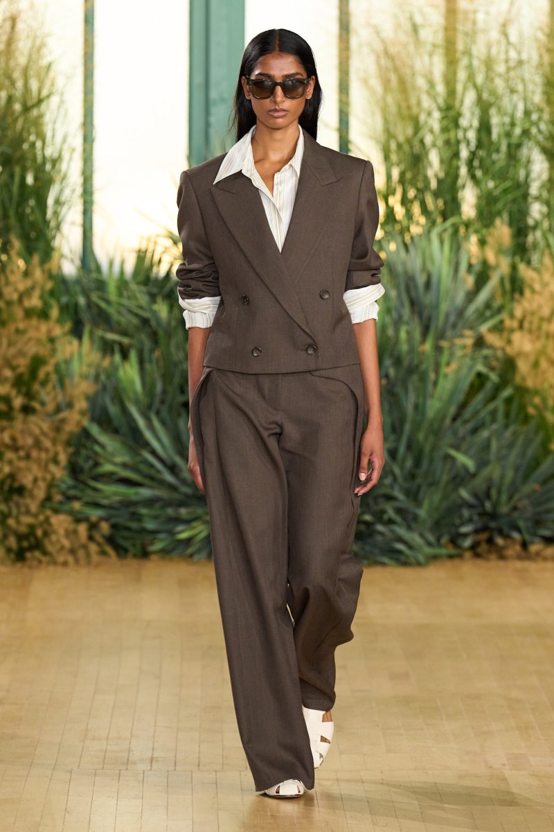 Model wearing a tailored brown double-breasted blazer with matching wide-leg trousers, white collared shirt, oversized sunglasses, and white open-toe heels in a runway setting