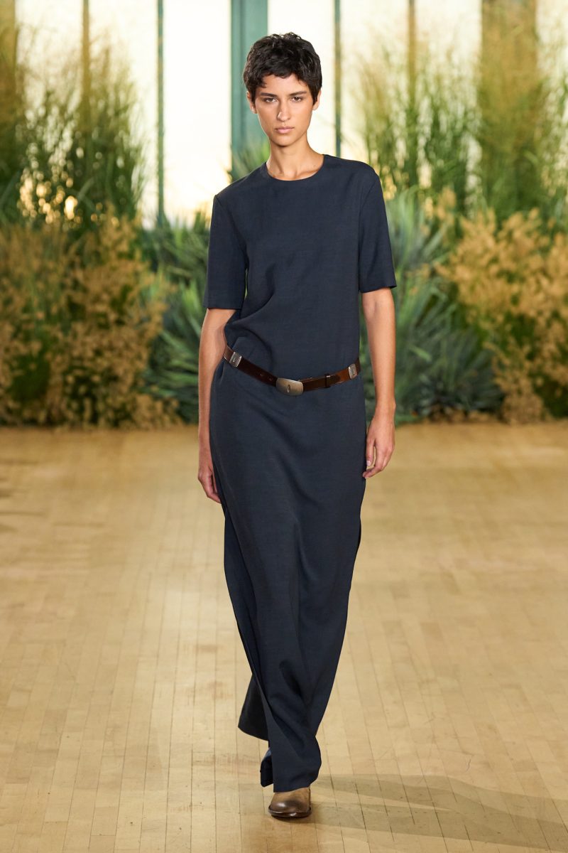 Model wearing a navy blue maxi dress with short sleeves and a brown leather belt, styled with brown ankle boots, runway setting with greenery background