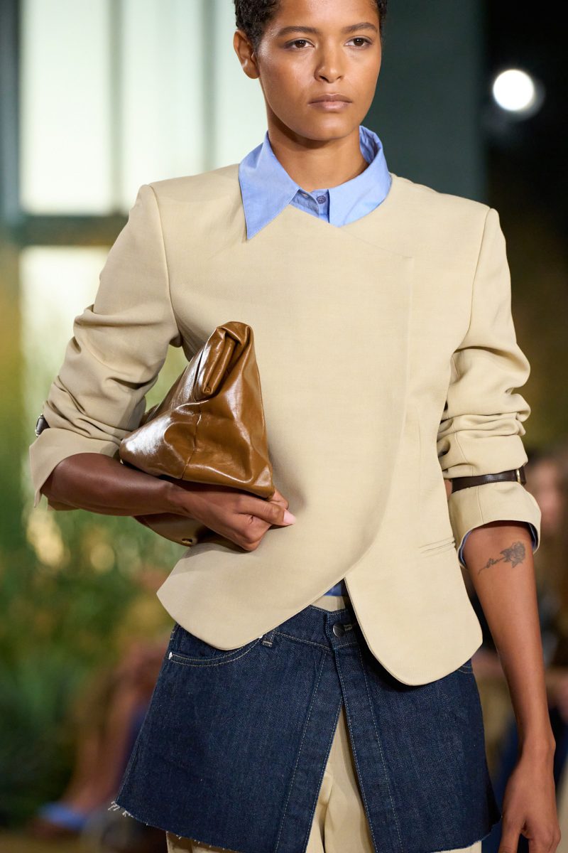 Model wearing a beige asymmetrical blazer over a blue collared shirt, paired with a dark denim skirt and holding a brown leather clutch bag