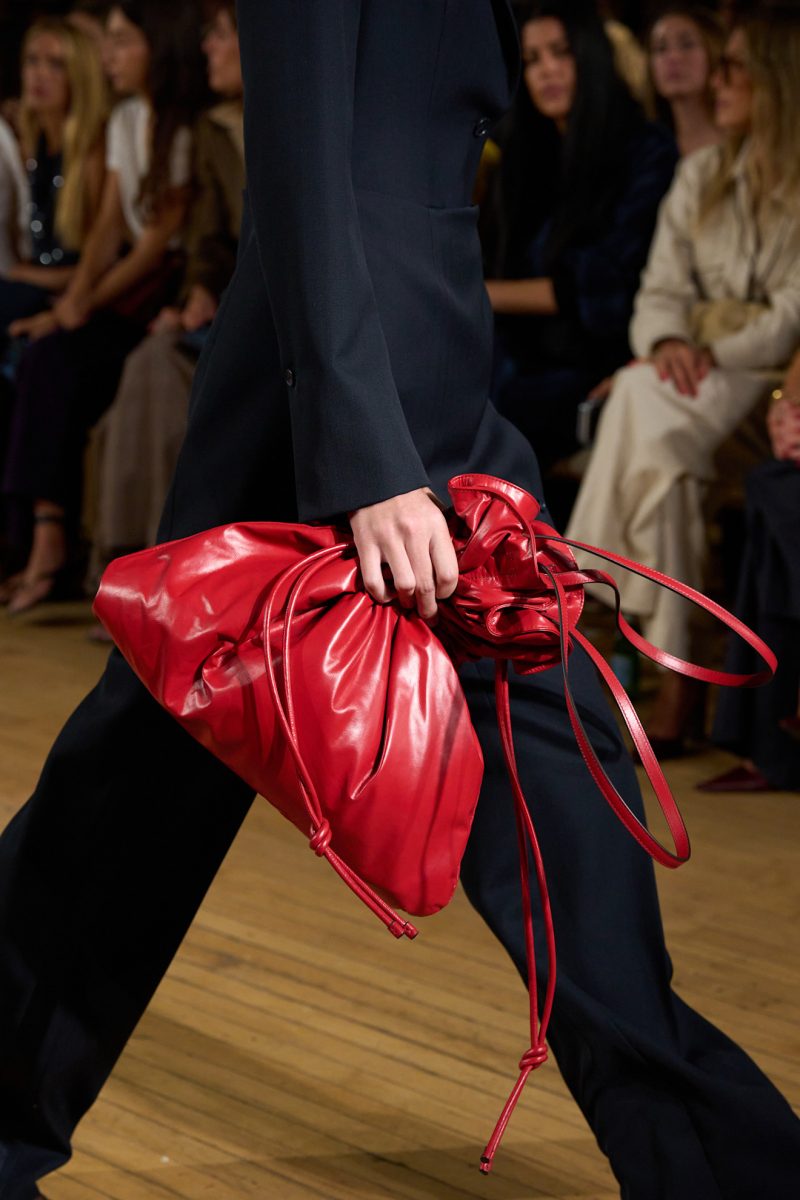 Model holding a bold red leather drawstring pouch bag paired with a sleek black tailored suit on the runway