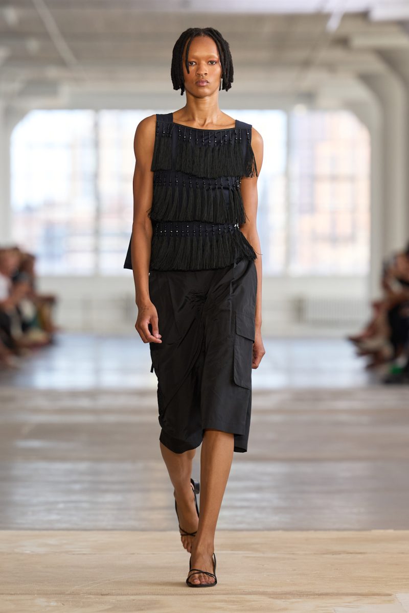 Model wearing a black sleeveless fringe top paired with black cargo-style knee-length shorts and strappy black heels on runway