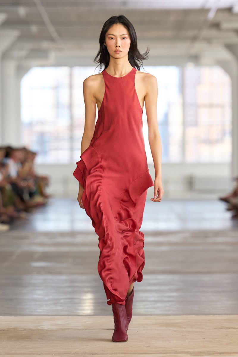 Model wearing a sleeveless red satin dress with ruffled details and matching red ankle boots on runway