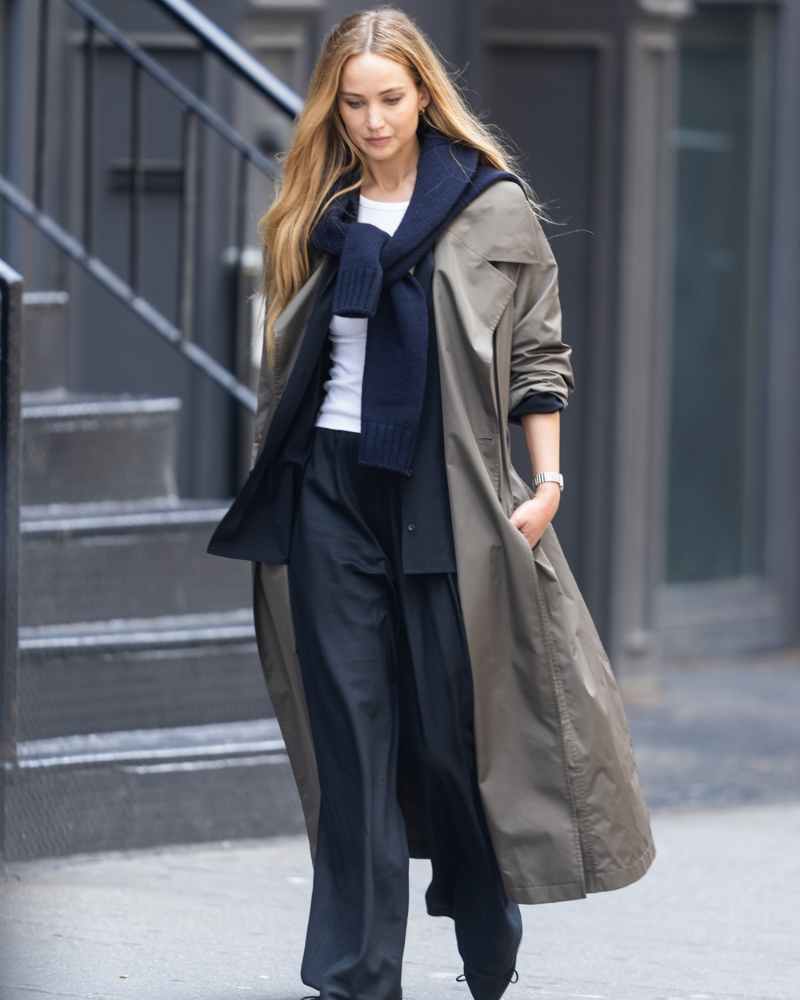 Model wearing a long olive trench coat over a white top and black wide-leg pants%2C styled with a navy sweater draped over shoulders