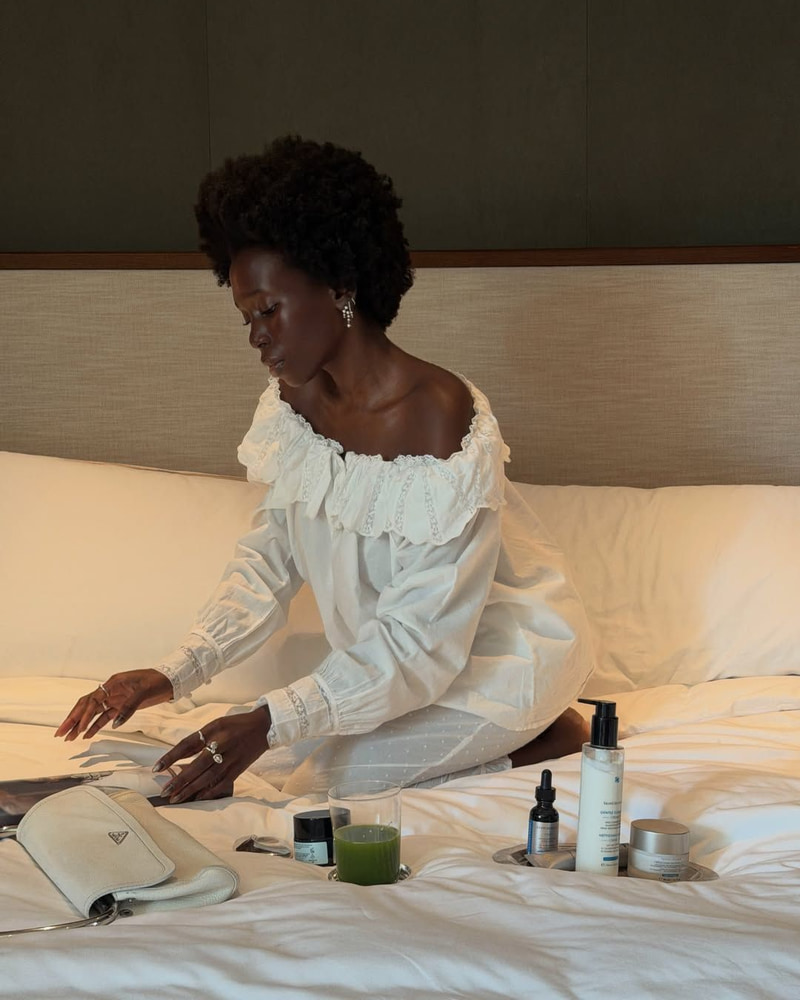 Elegant off-the-shoulder white blouse with lace detail%2C styled with a delicate ring%2C surrounded by skincare products and a green beverage on a cozy bed