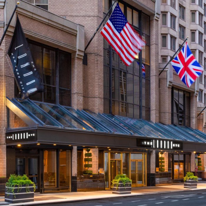 The lobby entrance of The London NYC hotel with glass canopy, American and British flags, and modern urban landscaping