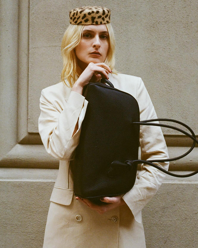 Blonde model in an ivory double-breasted blazer and leopard-print pillbox hat holding a black leather handbag against a stone wall
