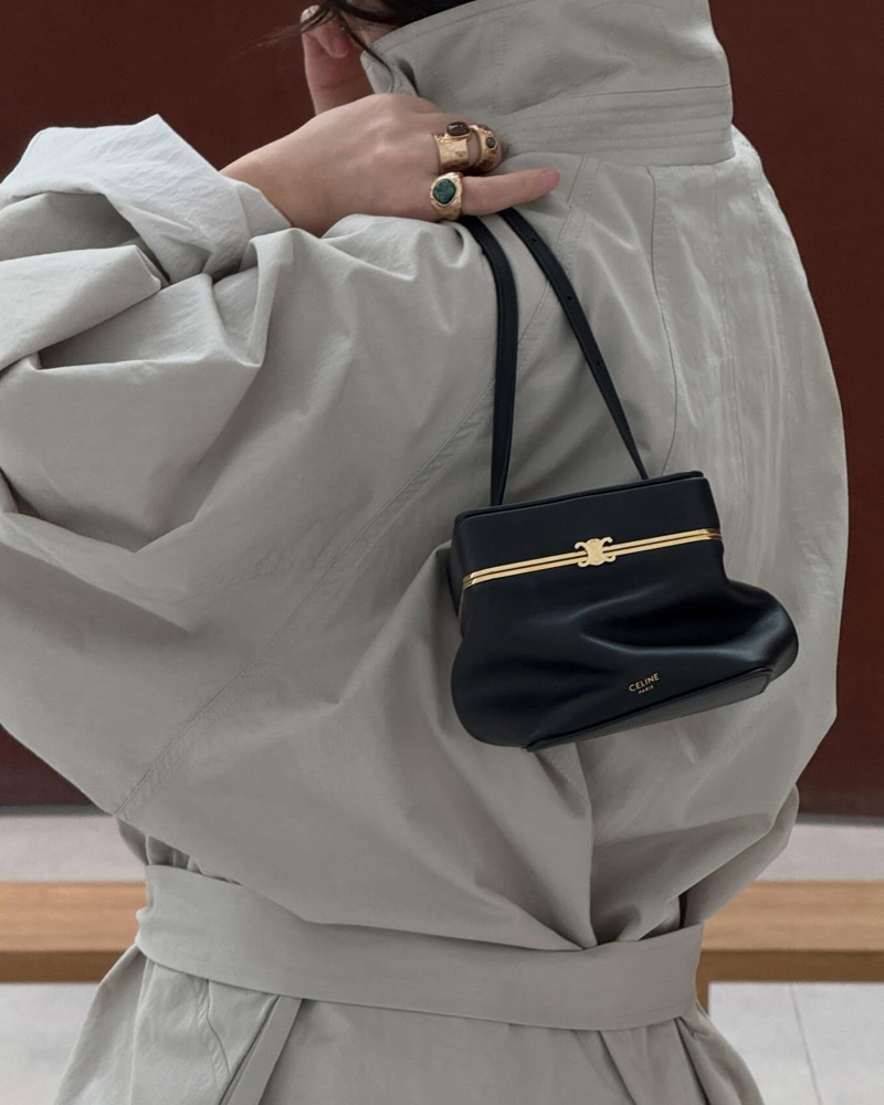 Celine black leather Cube bag with gold-tone hardware%2C styled with a light gray trench coat and statement rings