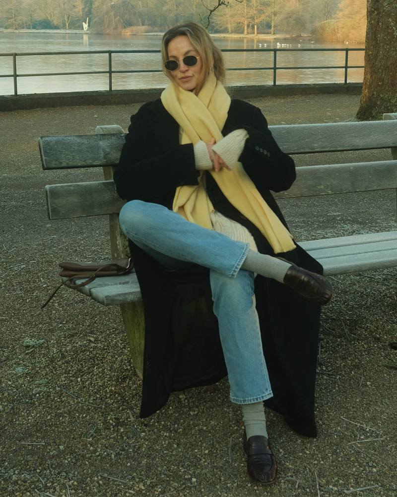Black coat styled with a yellow scarf%2C light-wash jeans and brown loafers while seated on a lakeside bench