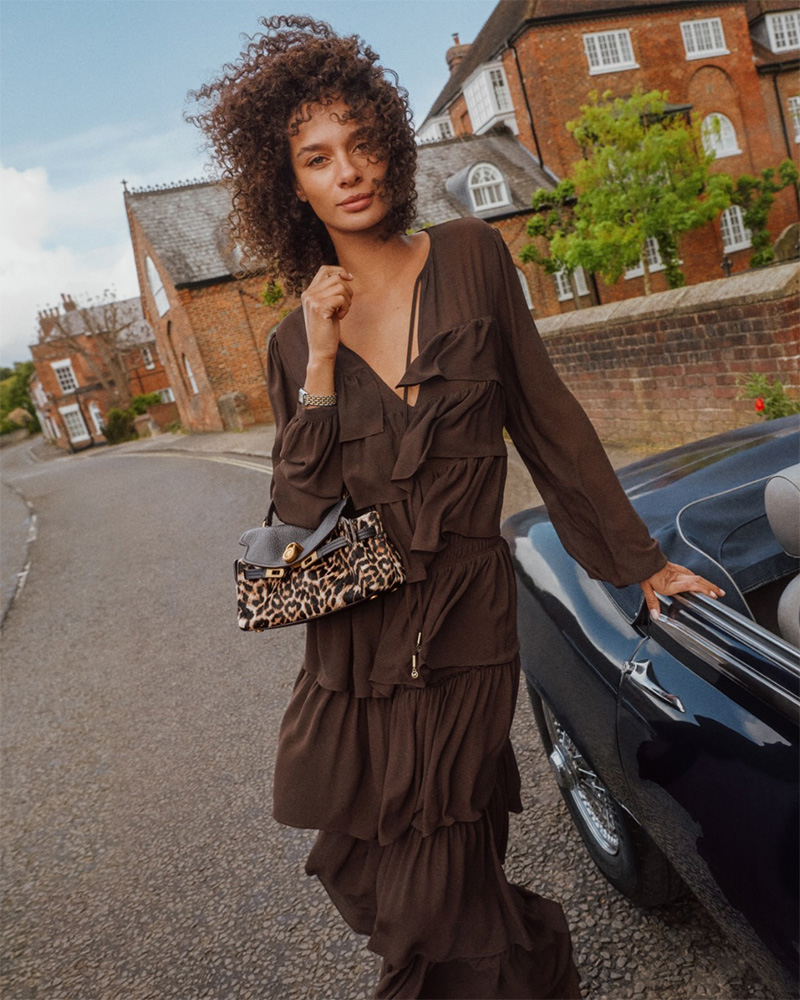 Brown tiered ruffle maxi dress styled with a leopard-print shoulder bag and gold watch beside a vintage car on a brick-lined street