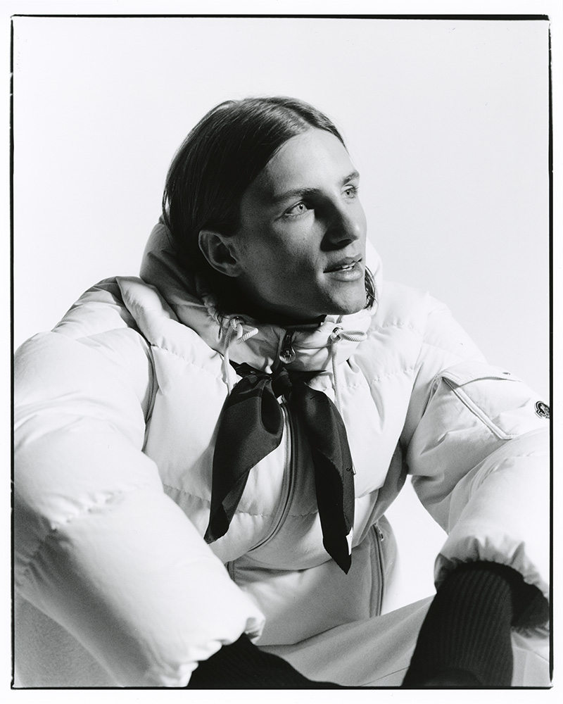 Black-and-white portrait of a model wearing a white Moncler hooded puffer jacket with a black scarf against a studio backdrop