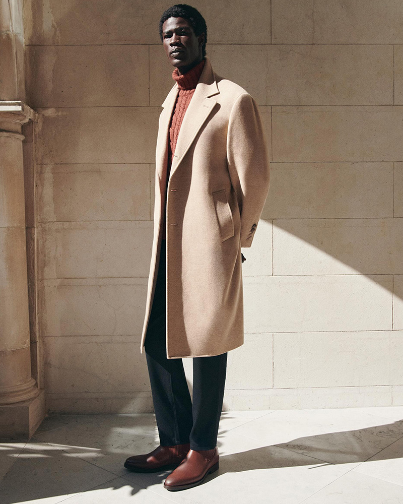Men%27s beige wool overcoat layered over a rust cable-knit turtleneck%2C dark trousers and brown leather boots in a sunlit hallway