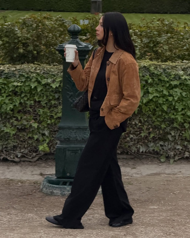 Brown suede jacket styled with black wide-leg trousers and boots while carrying coffee through a leafy park