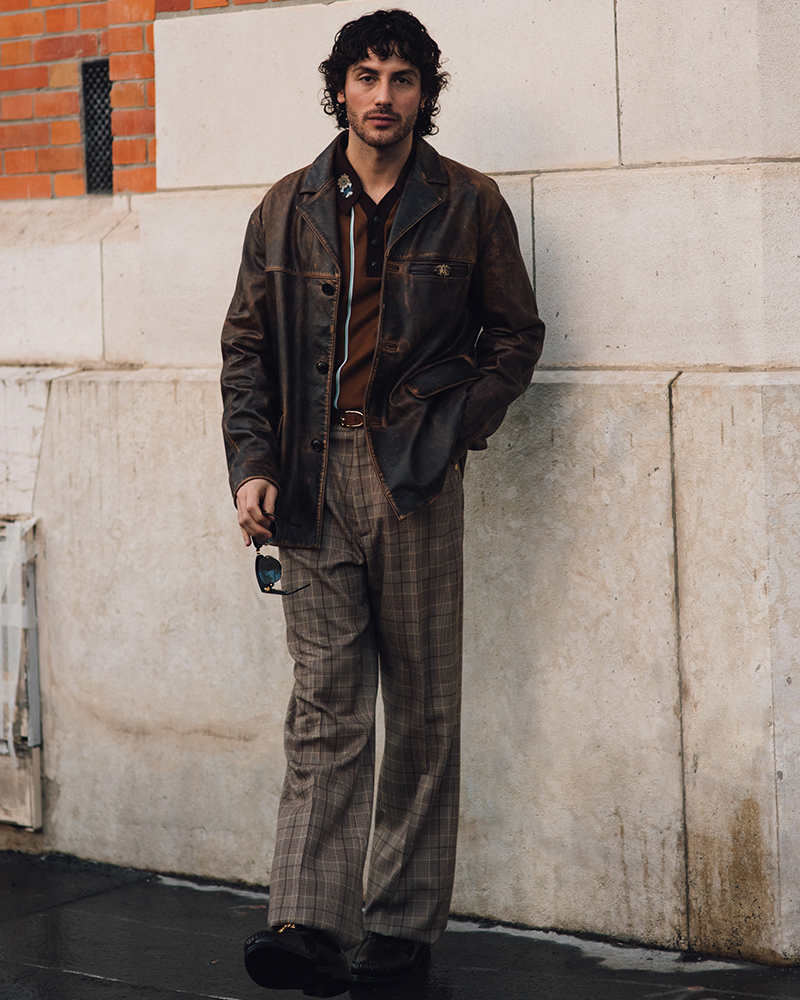 Brown leather jacket styled with plaid trousers%2C striped polo and black shoes against a stone city wall