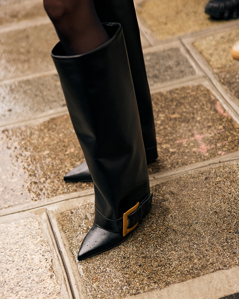 Black knee-high leather boots with pointed toes and oversized gold buckles%2C styled on wet pavement for a chic fall look