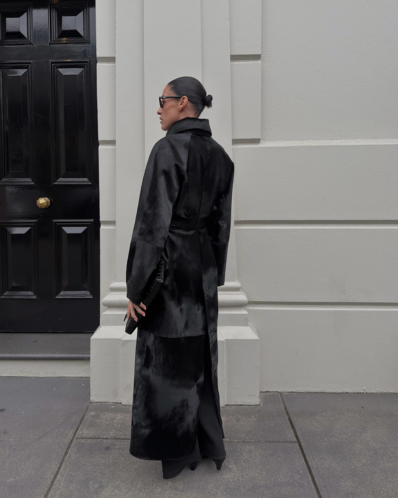 Street style look featuring a Nour Hammour black belted leather trench coat%2C sunglasses%2C and clutch against a white facade
