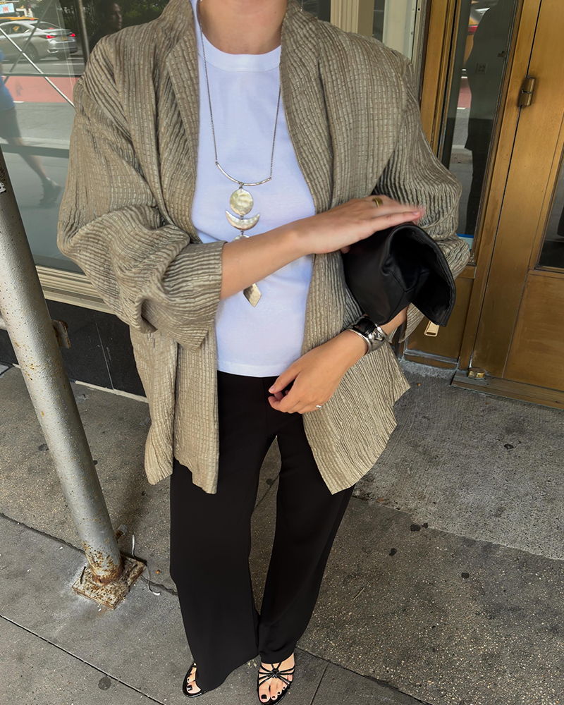 Chic outfit featuring a textured beige blazer%2C white T-shirt%2C black wide-leg trousers%2C statement necklace and clutch on a city sidewalk