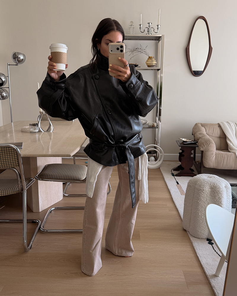 Woman wearing a belted black leather bomber jacket paired with beige wide-leg pants in a modern living room setting