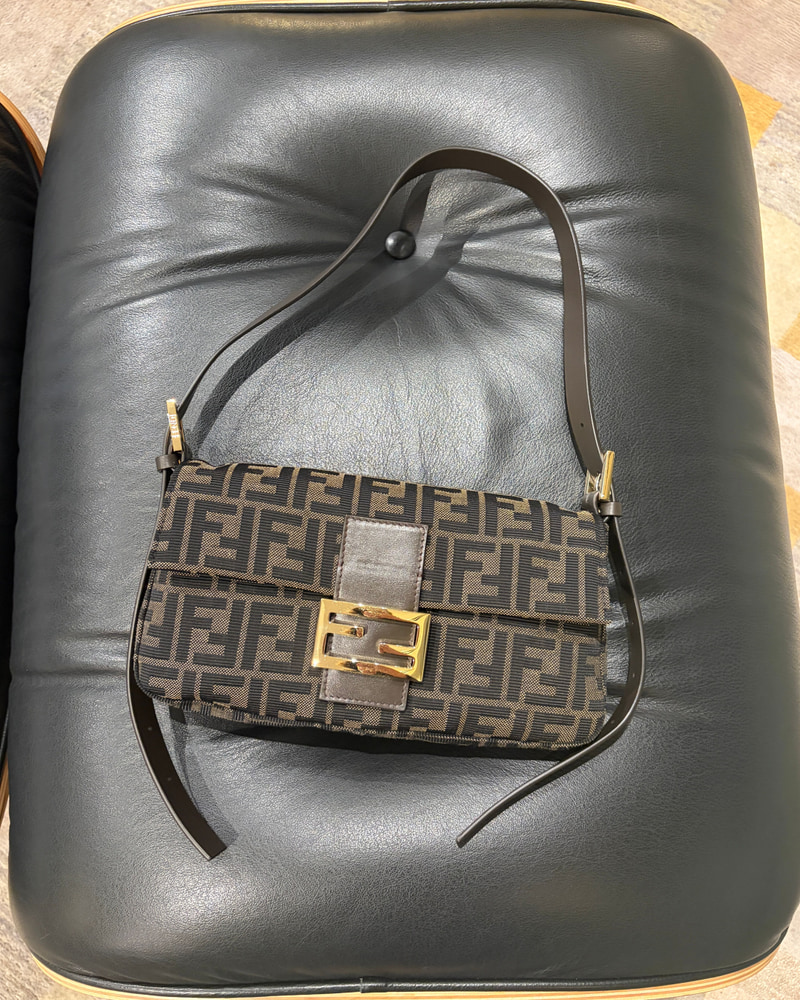 Fendi baguette bag in brown monogram canvas with gold-tone logo buckle and leather strap on black leather chair