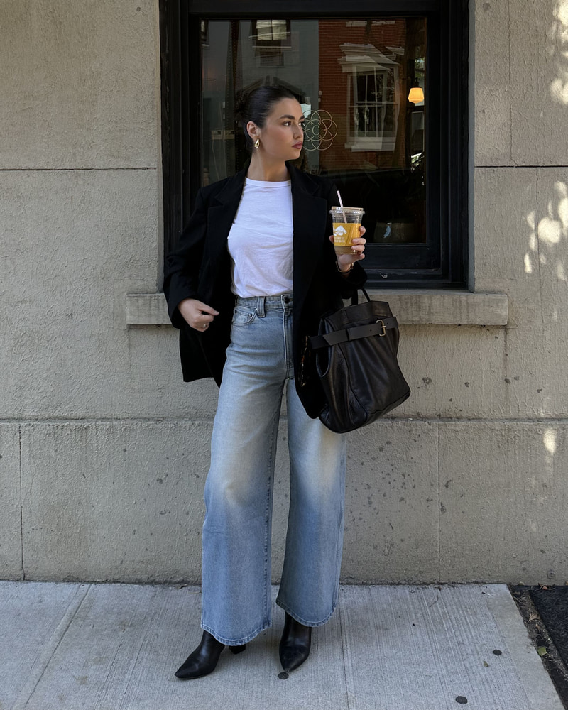 Woman wearing a black blazer%2C white t-shirt%2C wide-leg light wash jeans%2C black leather boots%2C and carrying a black leather handbag while holding a drink outdoors