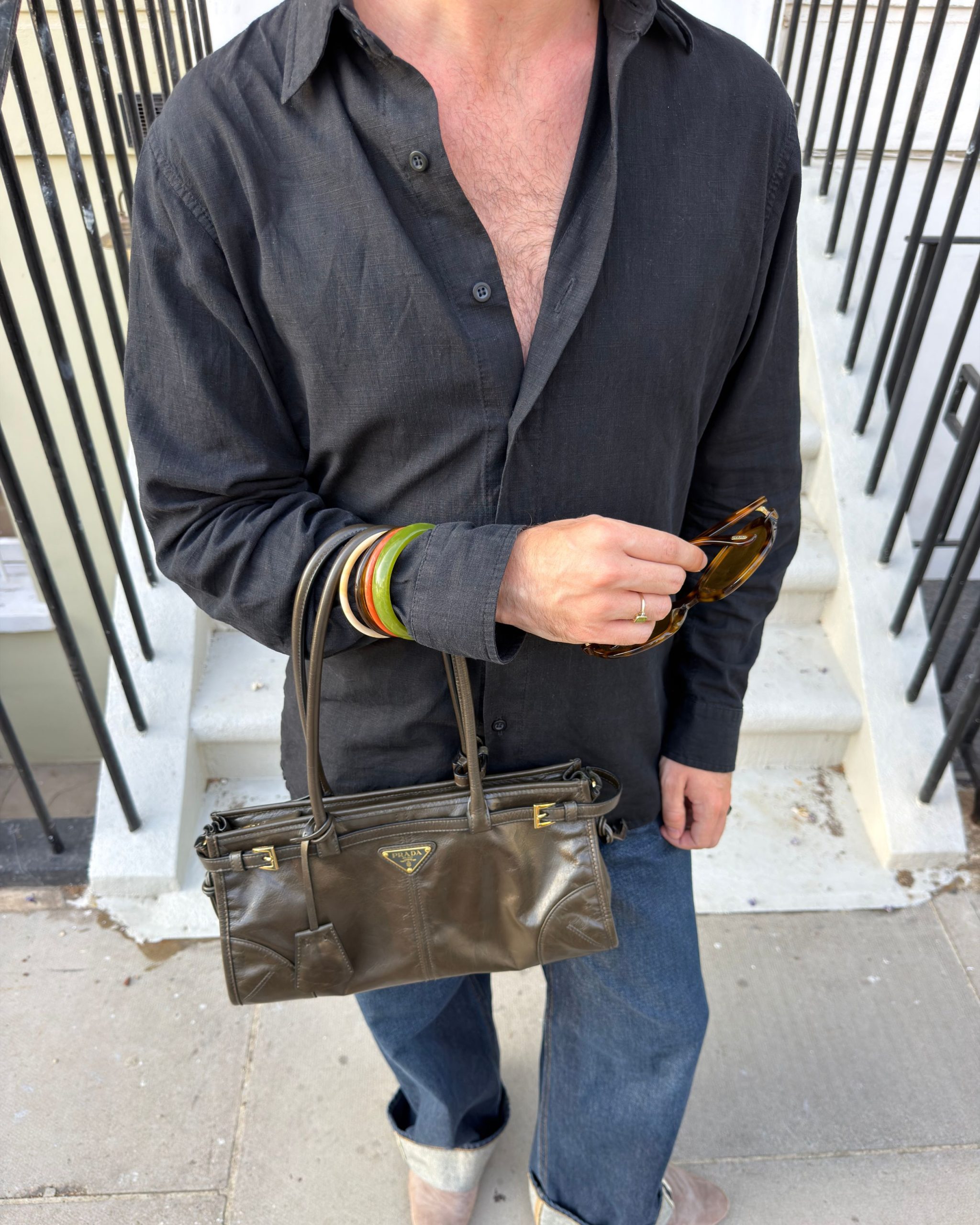 Man wearing a black linen shirt with blue cuffed jeans holding a brown Prada leather handbag and tortoiseshell sunglasses with colorful bangles on wrist