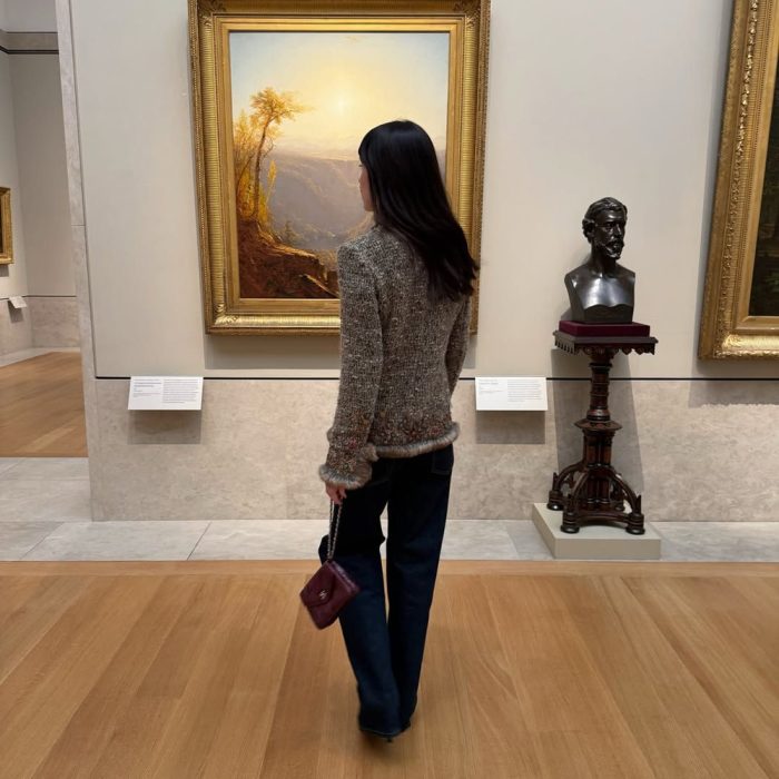 Chic autumn outfit featuring a cozy knitted sweater in earthy tones, high-waisted jeans, and a burgundy crossbody bag, set in an art gallery