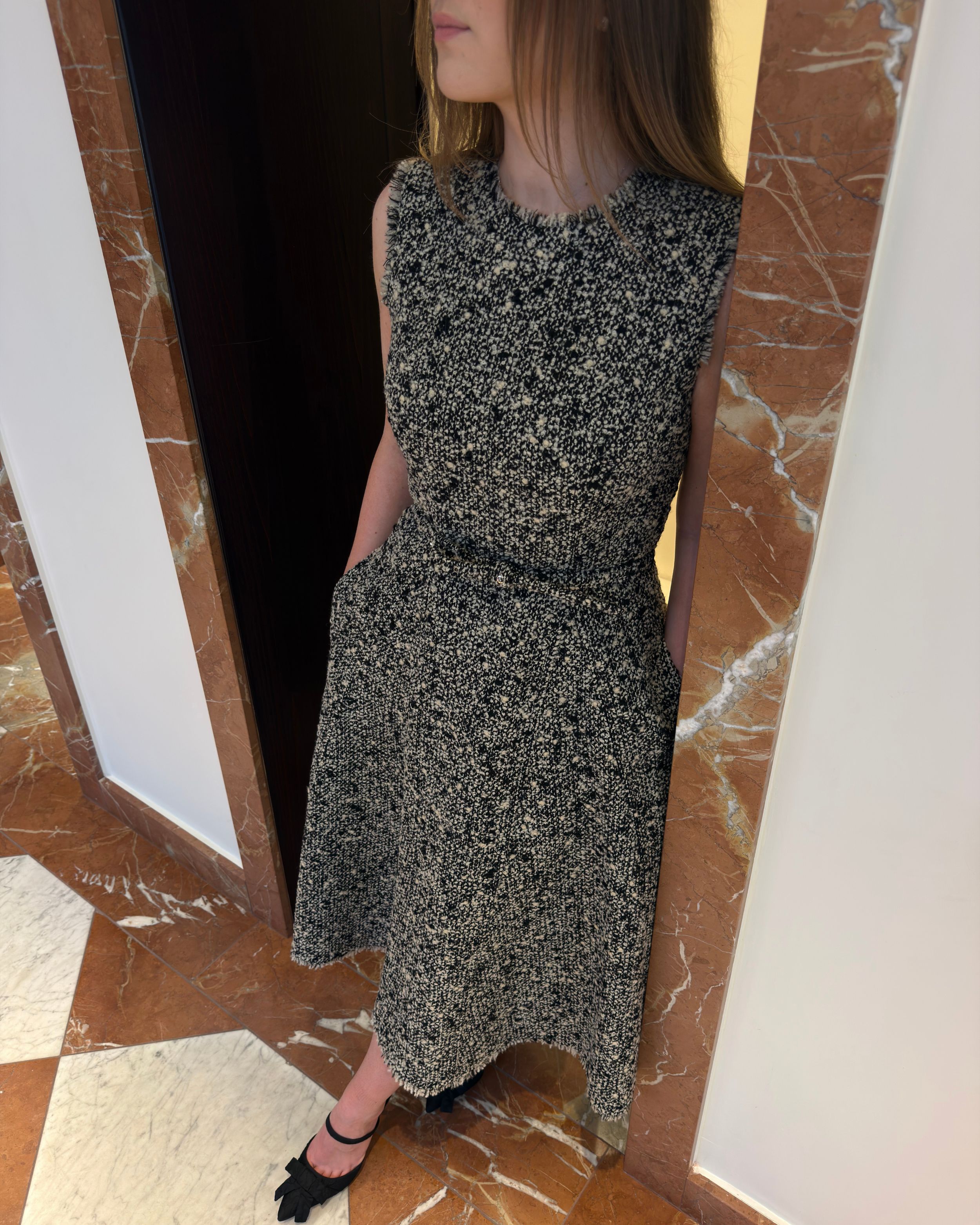 Sleeveless black and white tweed midi dress with pockets paired with black bow-embellished heels in a marble hallway setting